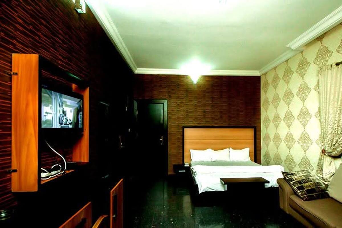 Quarto de hotel com parede de madeira, cama com roupa branca, televisão e sofá com almofadas estampadas.