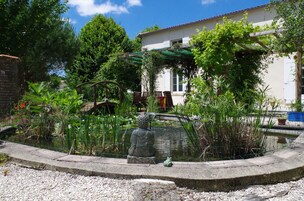 Um jardim tranquilo com um lago, uma estátua de Buda e uma casa ao fundo.