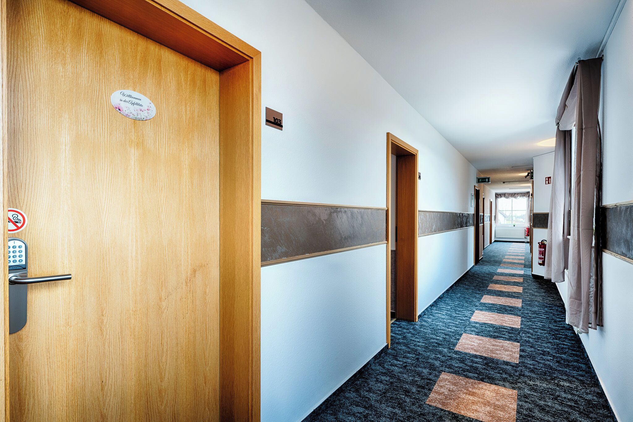 Um corredor de hotel com porta de madeira, fechadura eletrônica, placa com nome e tapete azul estampado.