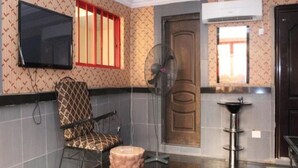 Quarto com televisão fixada na parede, uma cadeira estampada, um ventilador, uma porta e uma pequena mesa com pia.