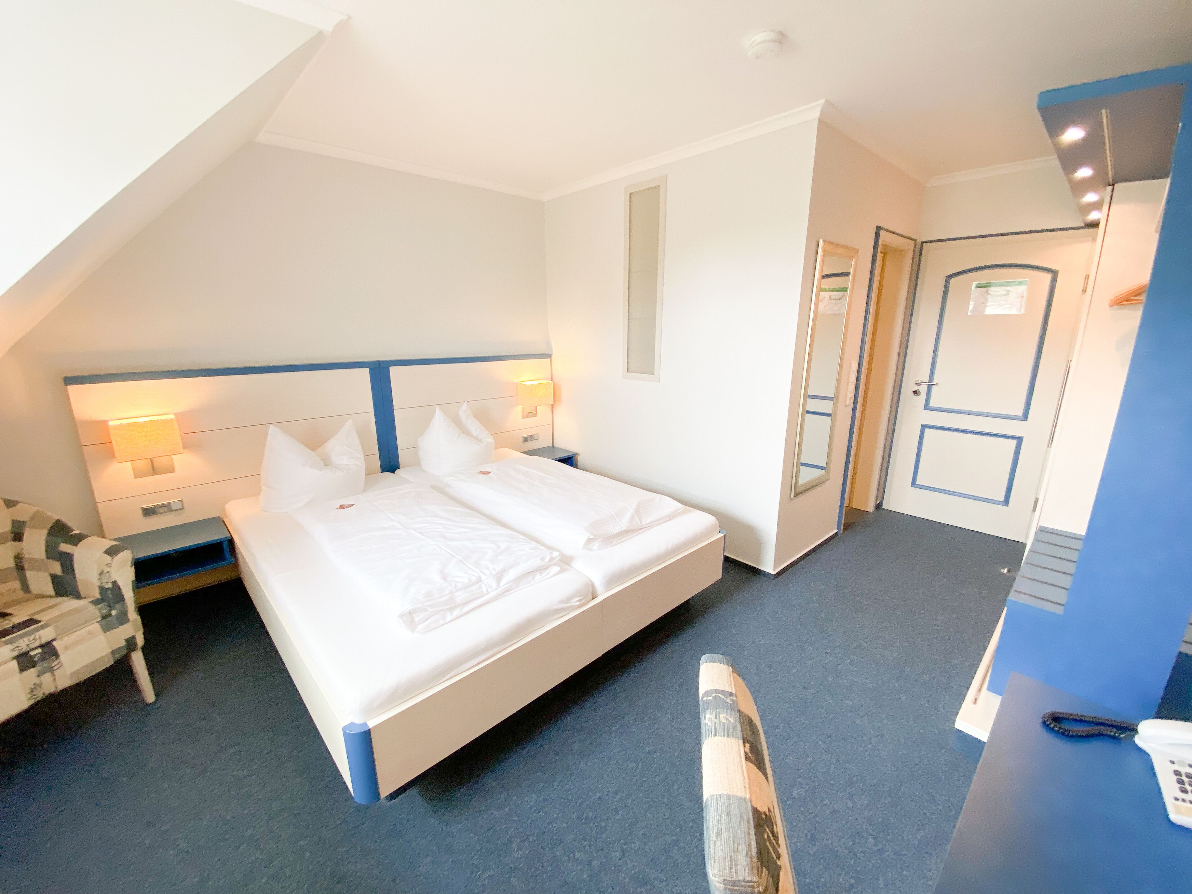 Quarto de hotel com uma cama grande, uma mesa e uma cadeira. Há um espelho na parede e uma porta com moldura azul.