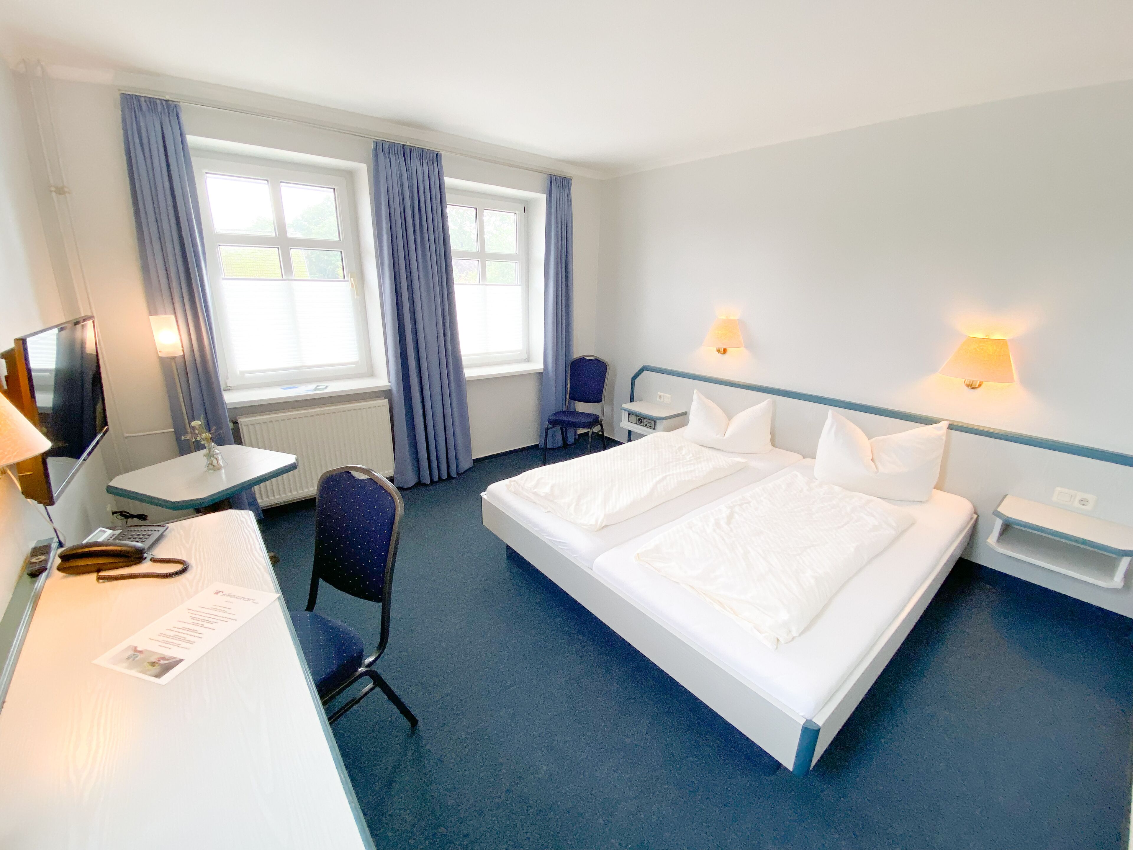 Quarto de hotel com uma cama grande, uma mesa com computador, uma cadeira e duas luminárias de cabeceira.