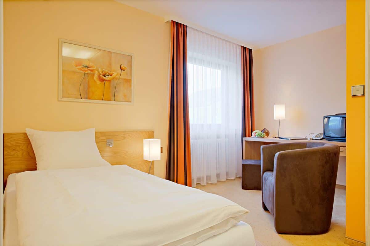 Quarto de hotel com cama, escrivaninha, cadeira, televisão e um quadro na parede.