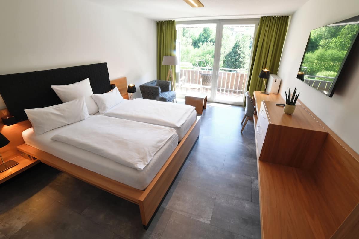 Quarto de hotel com cama de casal, escrivaninha com cadeira, televisão e varanda com vista para o verde.