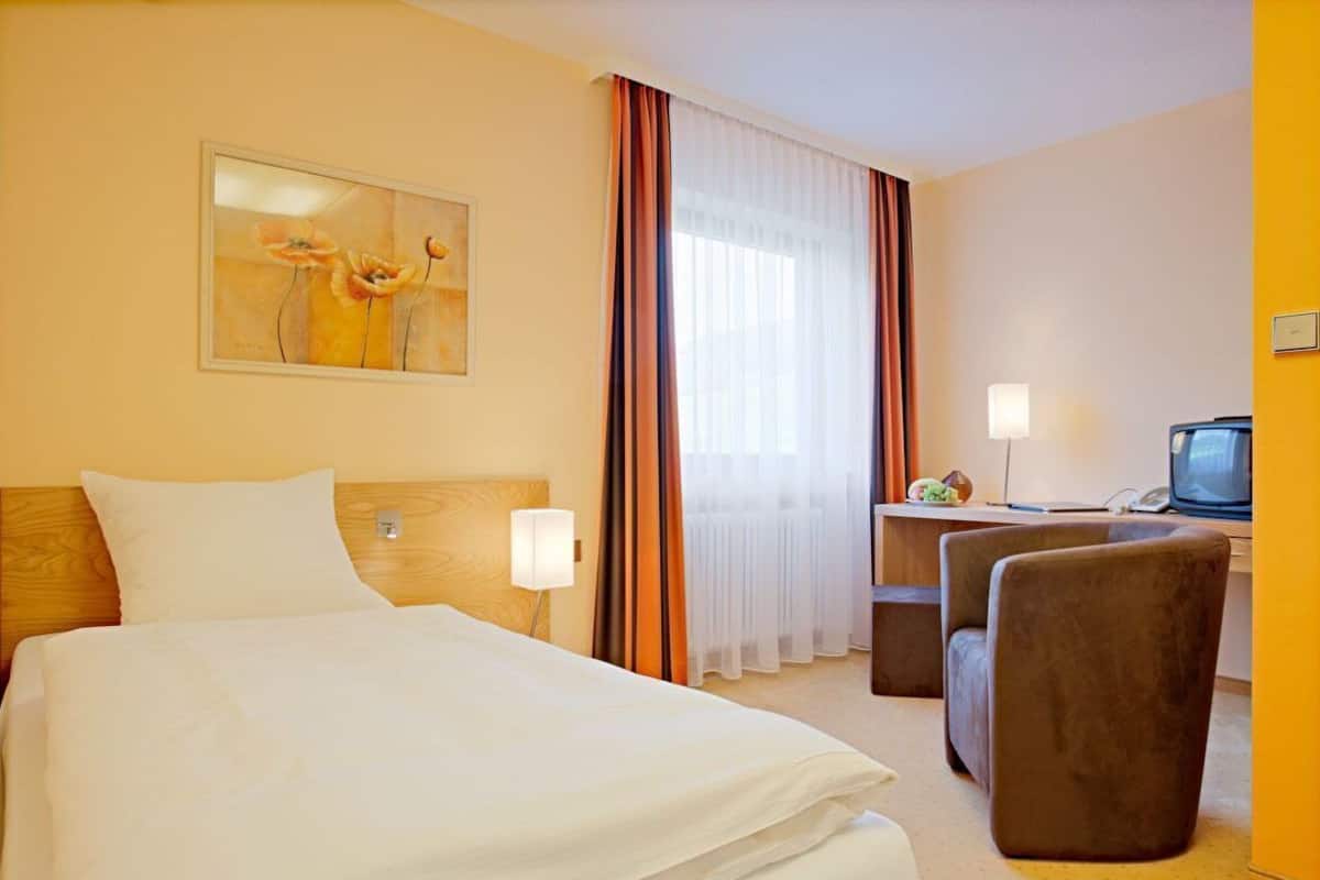 Quarto de hotel com cama, escrivaninha com televisão, cadeira e um quadro na parede.