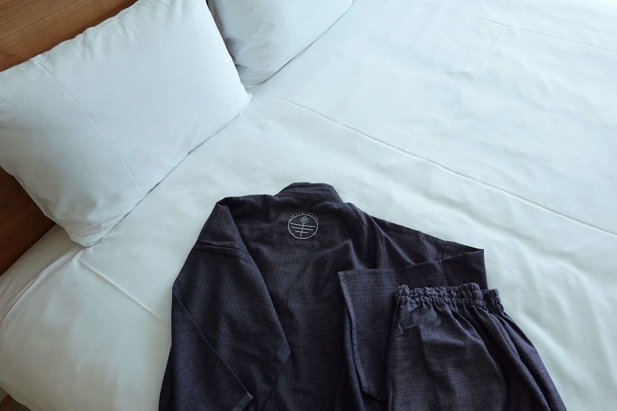 Cama bem arrumada com roupa de cama branca e um conjunto de roupa escura dobrado sobre ela.