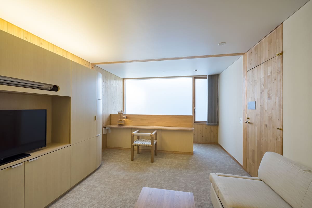 Quarto compacto com TV de tela plana, uma pequena escrivaninha de madeira, uma cadeira e um sofá.
