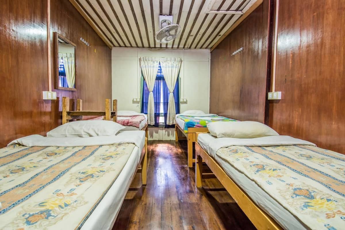 Quarto com duas camas de madeira, um espelho, um ventilador de teto e uma janela com cortinas.