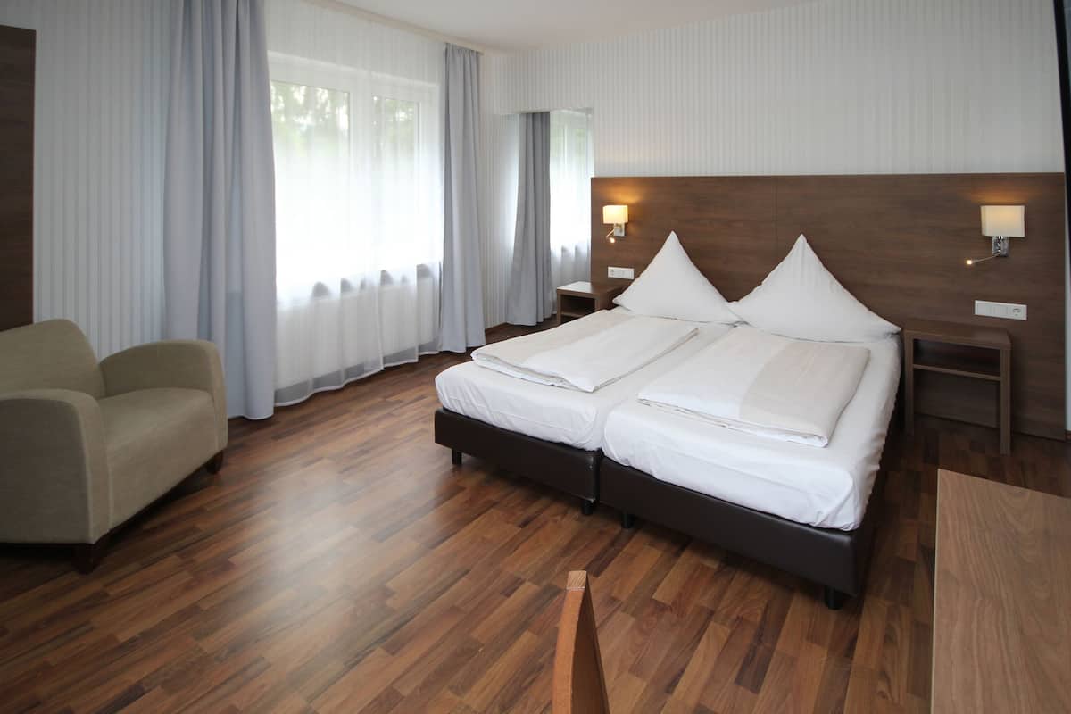 Uma cama de casal com cabeceira de madeira, uma mesa de cabeceira com luminária, um sofá e uma janela grande com cortinas.