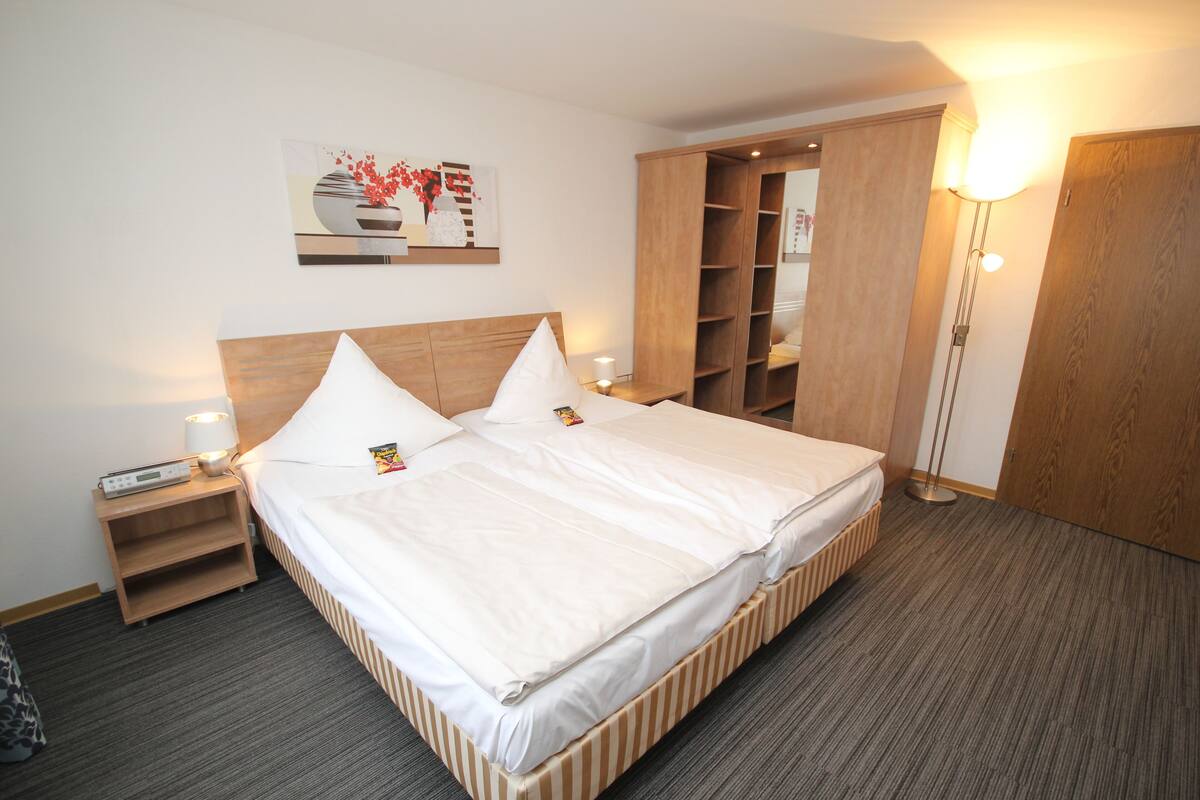 Quarto de hotel com uma cama grande, mesa de cabeceira com abajur, criado-mudo, guarda-roupa de madeira e um quadro na parede.