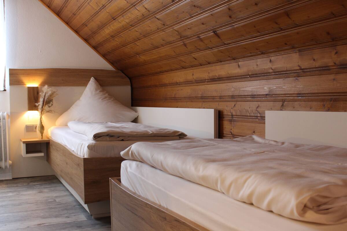 Quarto com teto inclinado, cabeceira de madeira e mesa de cabeceira com abajur.