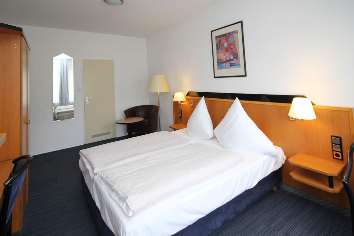 Quarto de hotel com uma cama grande, duas mesas de cabeceira com luminárias, uma cadeira, uma escrivaninha e um quadro na parede.