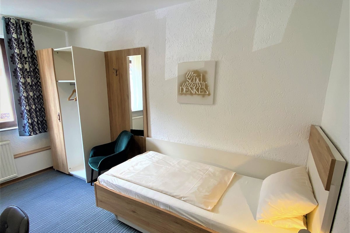 Quarto de hotel pequeno com uma cama de solteiro, uma escrivaninha, uma cadeira e um guarda-roupa.