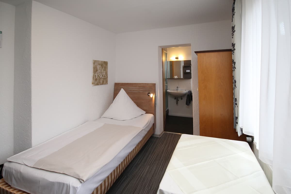 Quarto de hotel com uma cama de solteiro, uma mesinha de cabeceira, uma pequena mesa com uma placa, um espelho, uma pia e uma porta para o banheiro.