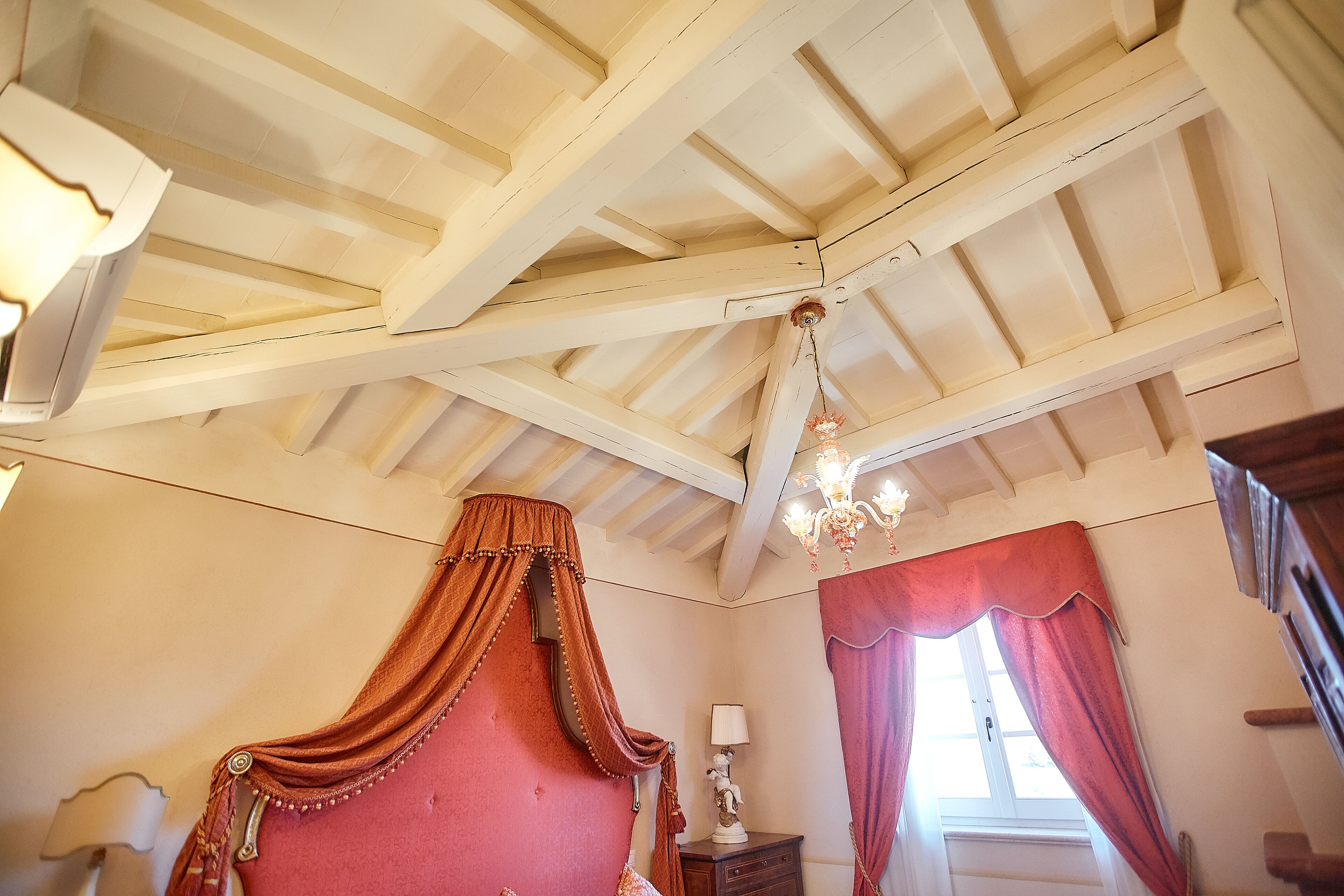 Quarto com teto alto, vigas de madeira, cama com dossel, lustre e janela com cortinas.
