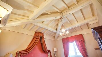 Quarto com teto alto, vigas de madeira, cama com dossel, lustre e janela com cortinas.