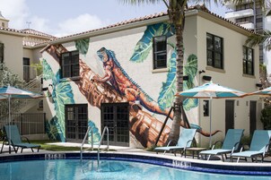 Uma piscina com espreguiçadeiras e guarda-sóis, um mural de um dinossauro, e um edifício com várias janelas e portas.
