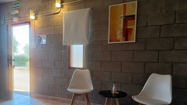 Quarto com uma porta identificada como 'SALIDA', duas cadeiras brancas, uma mesa redonda pequena e um quadro na parede.