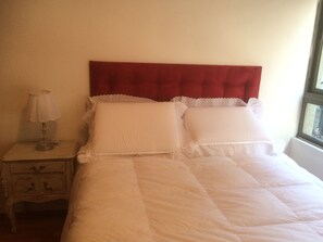 Uma cama bem arrumada com cabeceira vermelha, uma mesinha de cabeceira com abajur e uma janela com vista para o exterior.