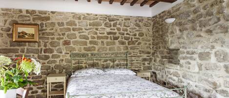 Quarto com parede de pedra, estrutura de cama de metal, mesa de cabeceira e criado-mudo.