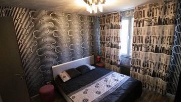 Quarto com cama, mesa de cabeceira, um banquinho vermelho, papel de parede estampado e cortinas com texto.