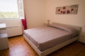 Quarto com cama, mesa de cabeceira, luminária e janela com vista para vegetação.