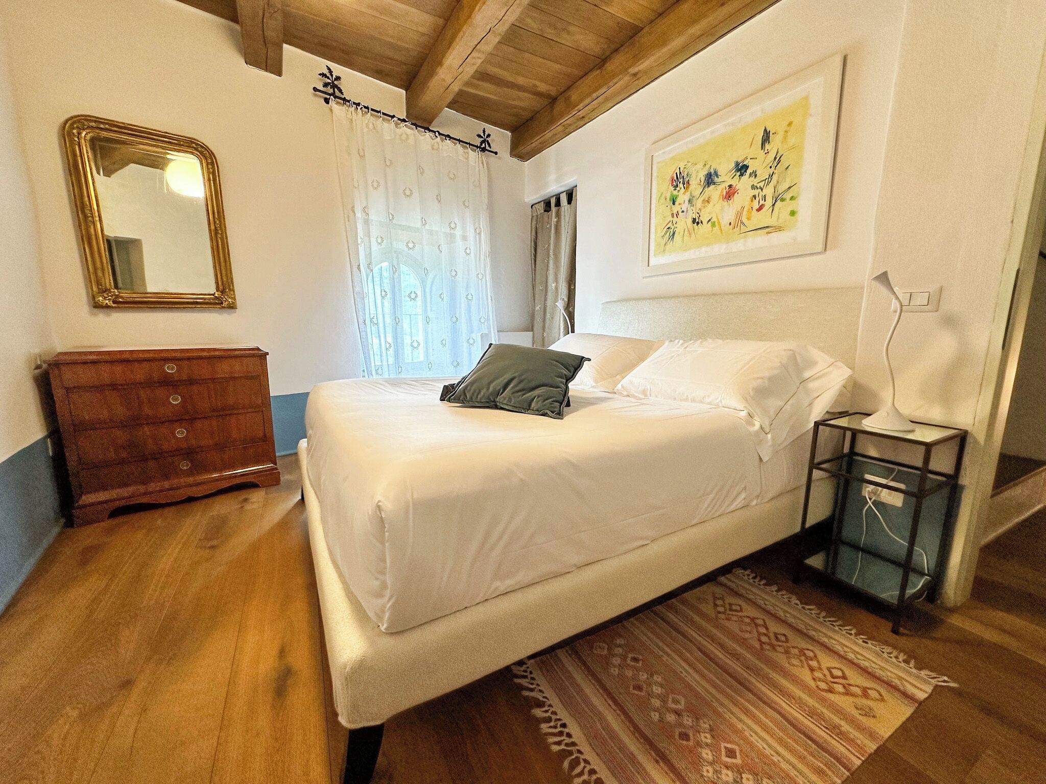 Quarto com cama, cómoda, mesa de cabeceira, luminária, espelho e um quadro na parede.