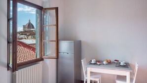 Um quarto pequeno com janela, uma mesa com cadeiras, um refrigerador e vista para um edifício com cúpula.