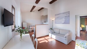 Uma sala de estar moderna com um sofá branco, uma mesa de jantar com cadeiras, uma televisão de tela plana na parede e uma área de cozinha ao fundo.