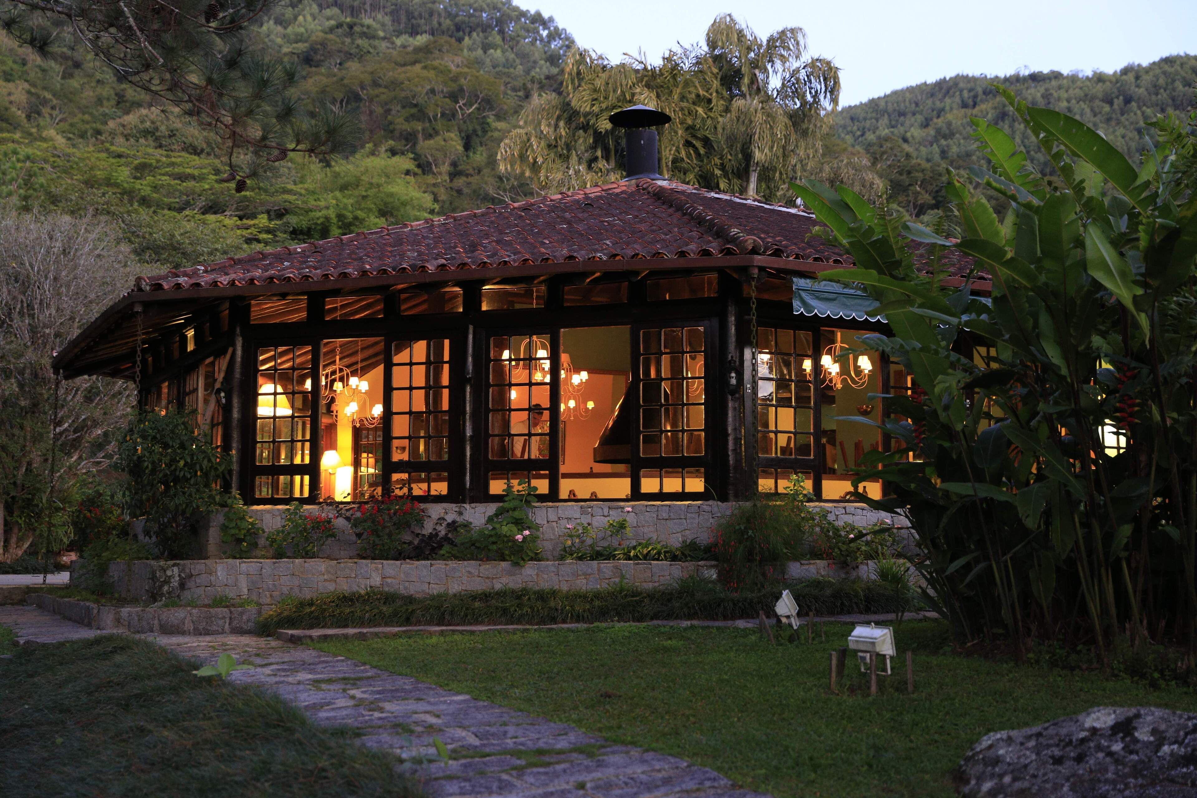 Uma casa de madeira rústica com janelas amplas, rodeada por vegetação exuberante e um caminho de pedra que leva à entrada.