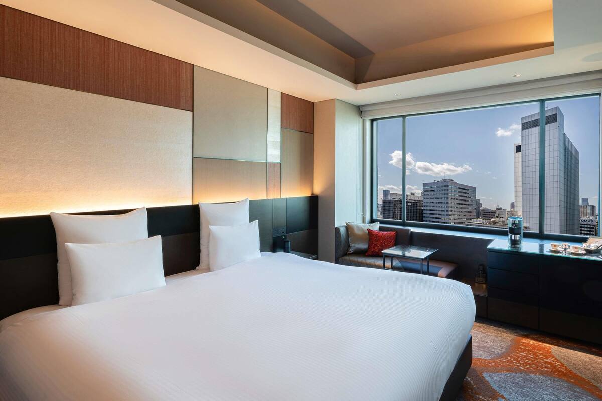 Quarto de hotel com uma cama grande, uma mesinha, uma cadeira e vista para a paisagem urbana através de uma janela ampla.