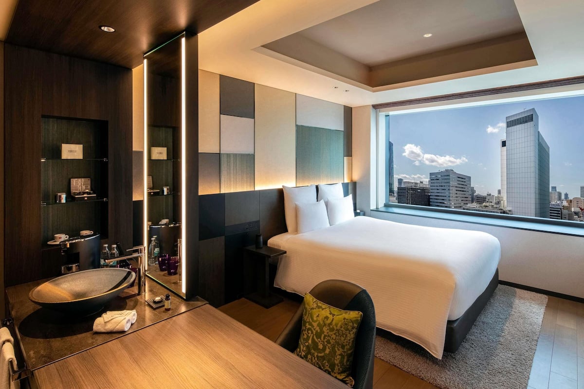 Quarto de hotel moderno com uma cama grande, vista para o horizonte da cidade, penteadeira com espelho e área de estar com mesa e cadeiras.