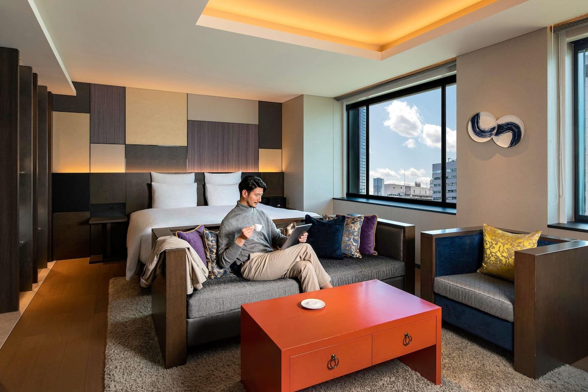 Quarto de hotel moderno com cama, sofá, mesa de centro e uma janela grande com vista para a cidade.