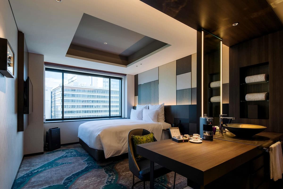 Quarto de hotel moderno com uma cama grande, uma escrivaninha com cafeteira, uma janela ampla com vista para a cidade e um banheiro com lavatório espaçoso e porta-toalhas.