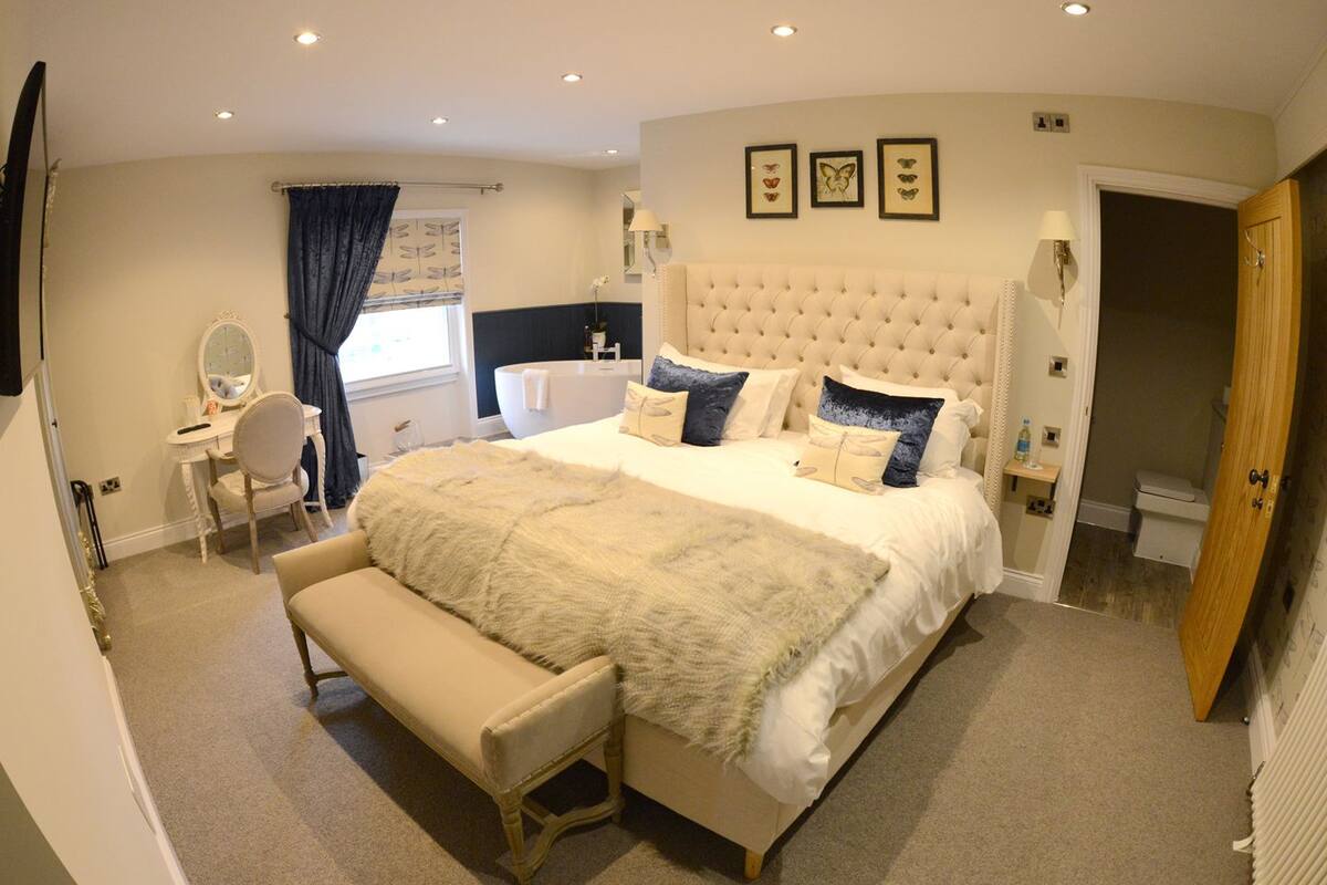 Um quarto espaçoso com uma cama grande, um banquinho, uma penteadeira com espelho, uma banheira e uma porta que dá para outro espaço.