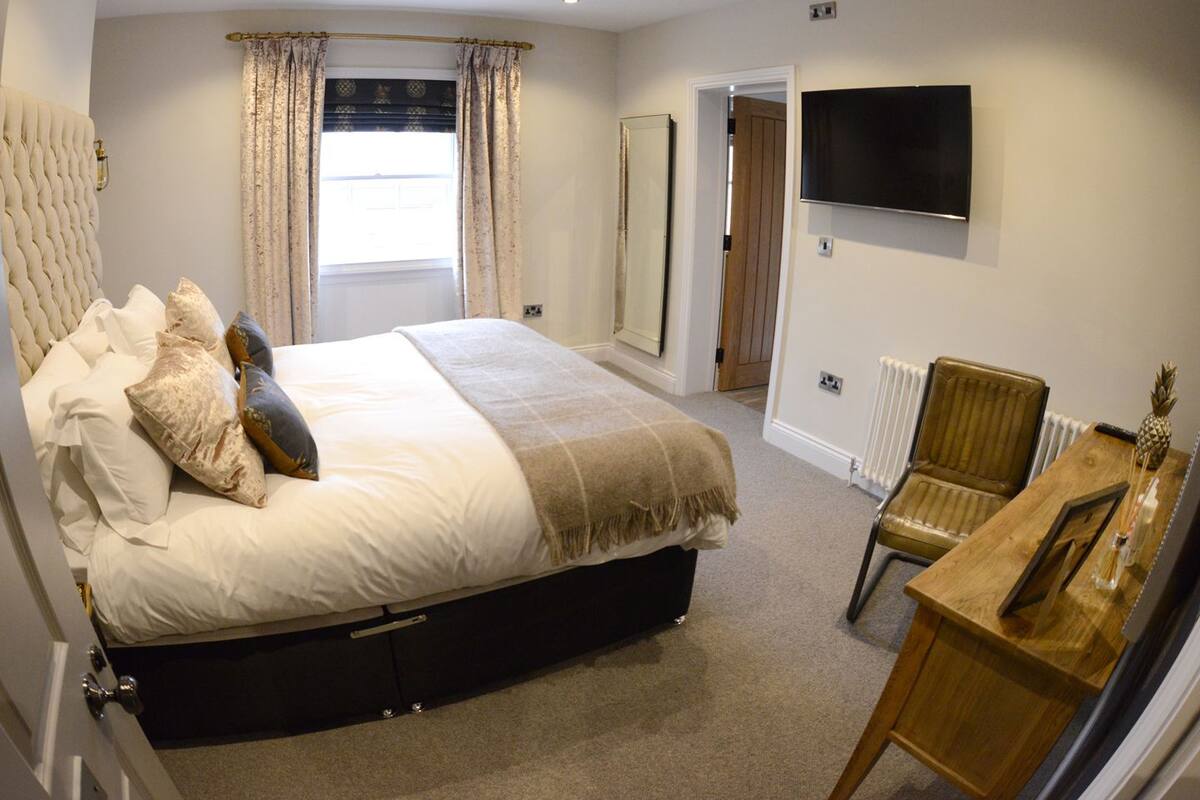 Quarto com uma cama grande, televisão fixada na parede, uma cadeira de madeira e um radiador.