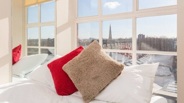 Quarto de hotel com uma janela ampla que oferece vista para a cidade, cama com roupa branca e duas almofadas, uma vermelha e outra bege.