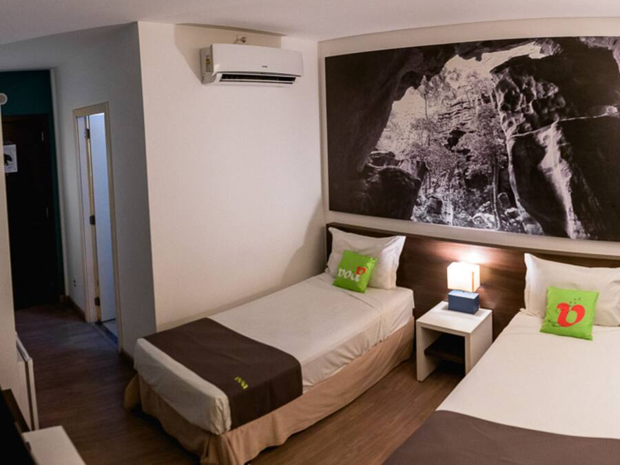 Quarto de hotel com duas camas de solteiro, mesa de cabeceira com abajur, um quadro grande emoldurado na parede e um aparelho de ar condicionado.