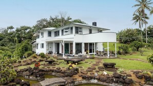 Uma casa branca de dois andares com varanda coberta e lago com pedras e plantas.