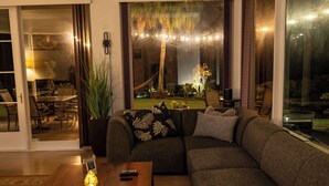 Uma sala de estar moderna com um sofá seccional, uma mesa de centro de madeira e vista para um jardim externo com luzes de festa.