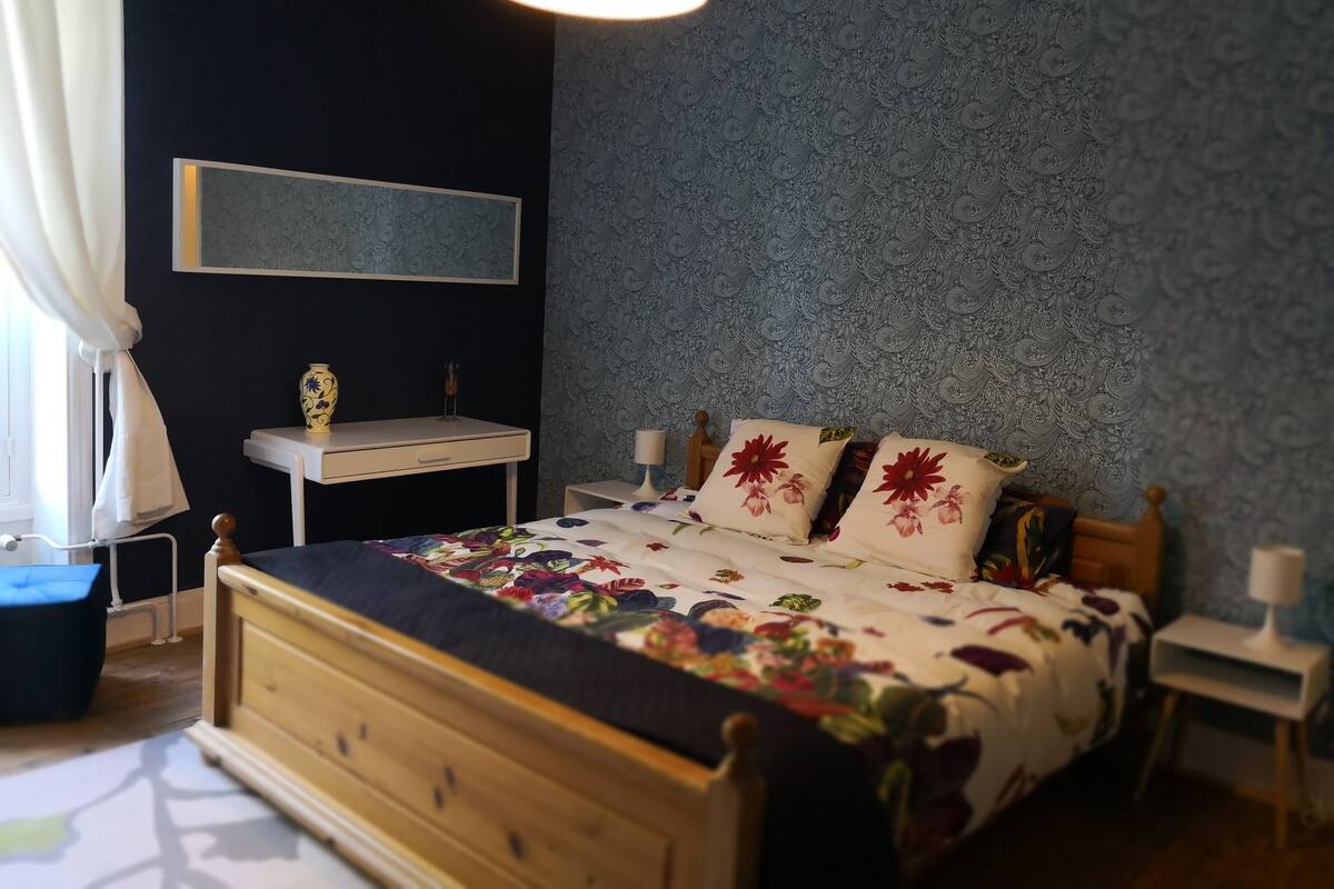 Quarto com cama, mesas de cabeceira, cômoda, espelho e janela com cortinas.