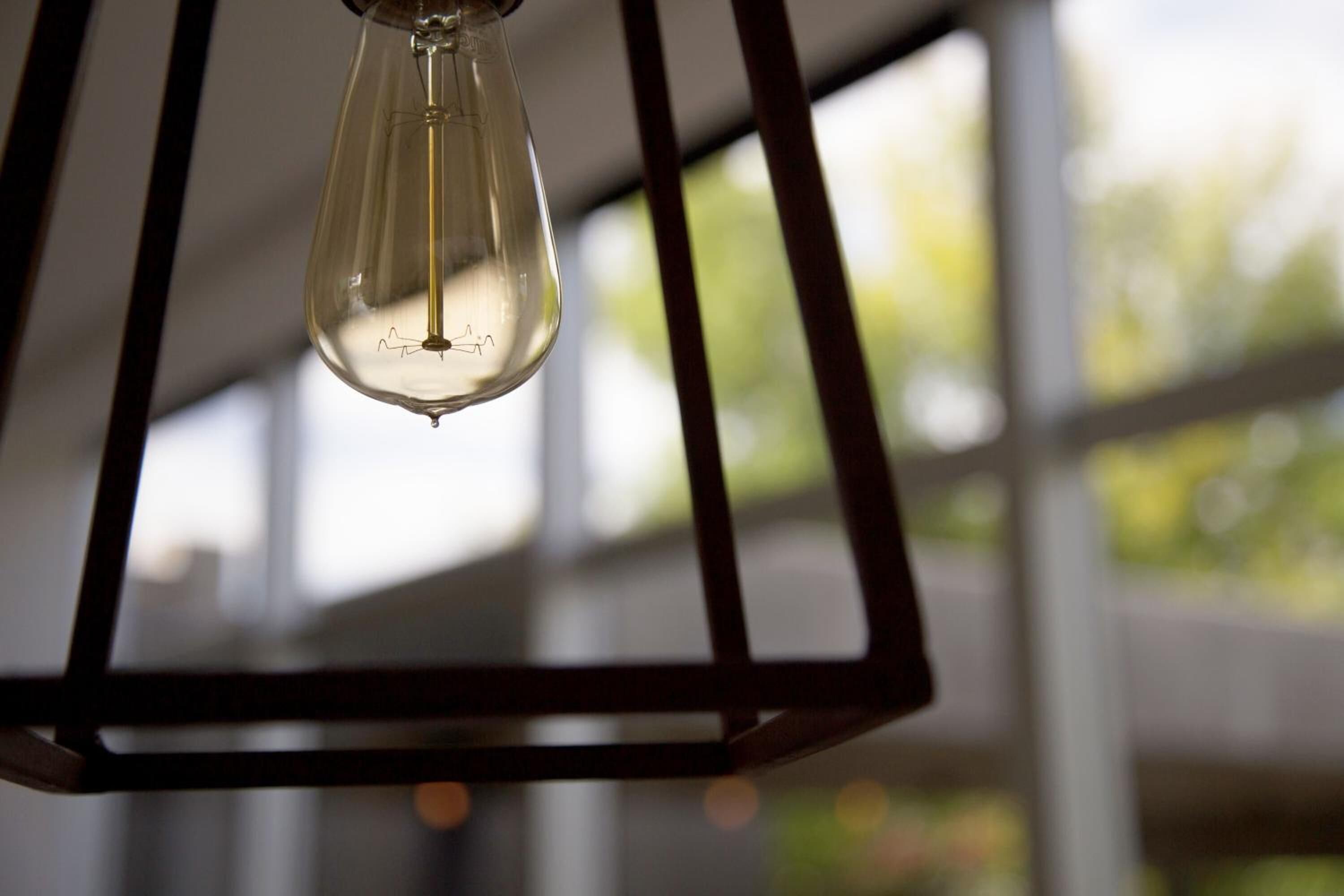 Luminária pendente com lâmpada de vidro transparente, suspensa por uma estrutura metálica geométrica.