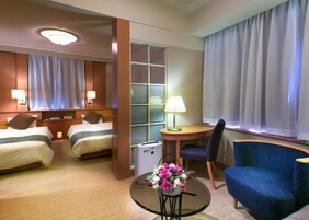 Quarto de hotel com duas camas, uma escrivaninha com abajur, uma cadeira e uma pequena mesa com flores.