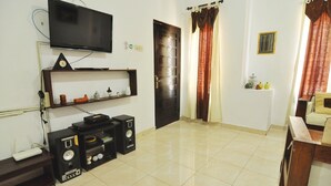 Uma sala de estar com televisão na parede, uma porta, janela com cortinas e um pequeno aparador com objetos decorativos.