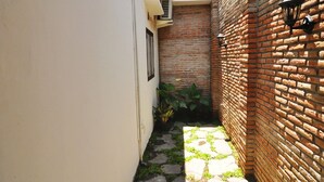 Um beco estreito com uma parede de tijolos de um lado e uma parede branca do outro. Há uma pequena área de jardim com pedras e plantas.