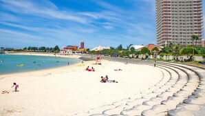 Uma praia com uma passarela de pedra curva, um hotel alto e uma orla de areia com pessoas e guarda-sóis.