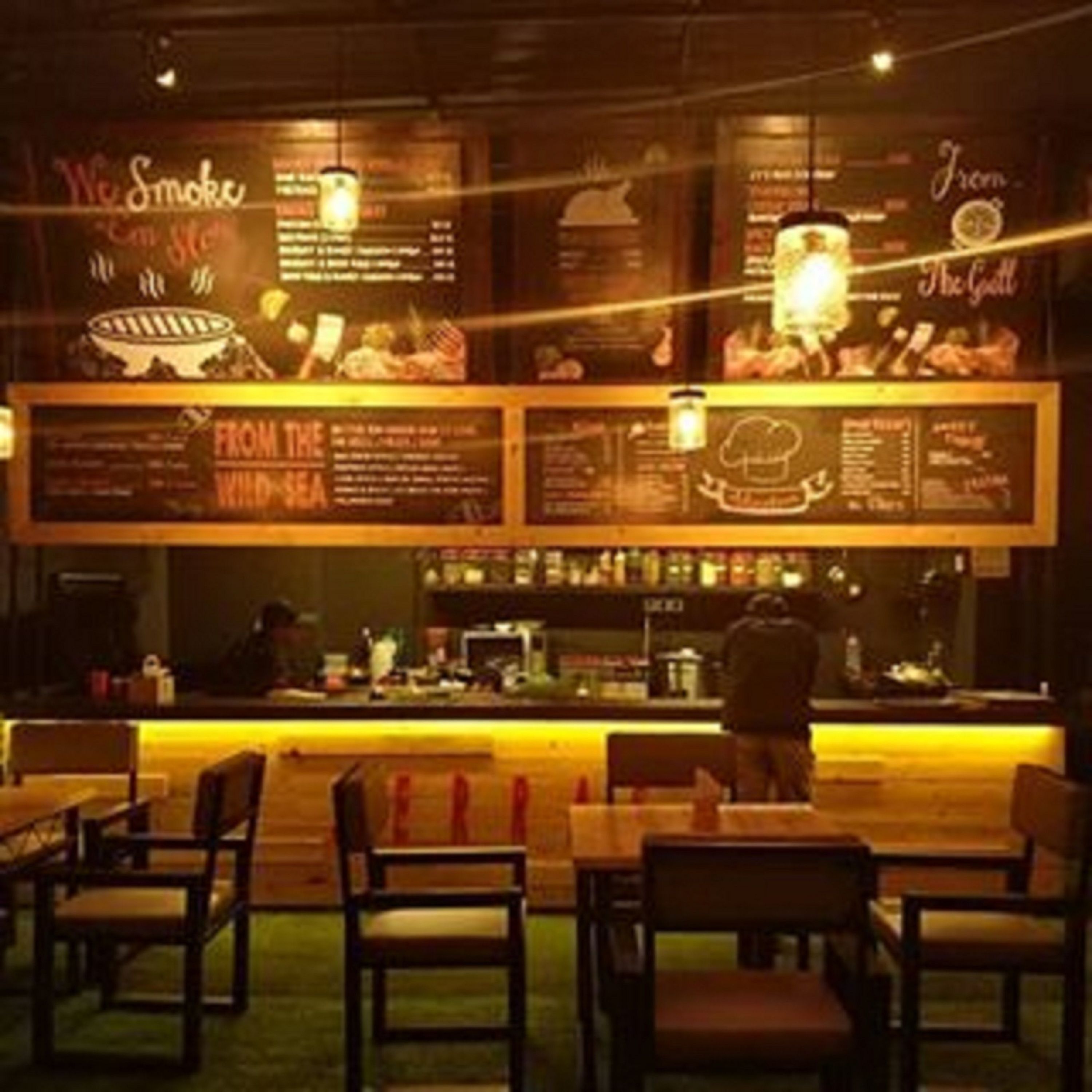 Interior de restaurante com área de bar, mesas e cadeiras de madeira e quadros de menu na parede.