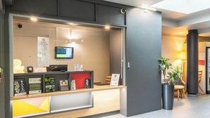 Uma mesa de recepção com tela digital, cartazes informativos e um vaso de planta.
