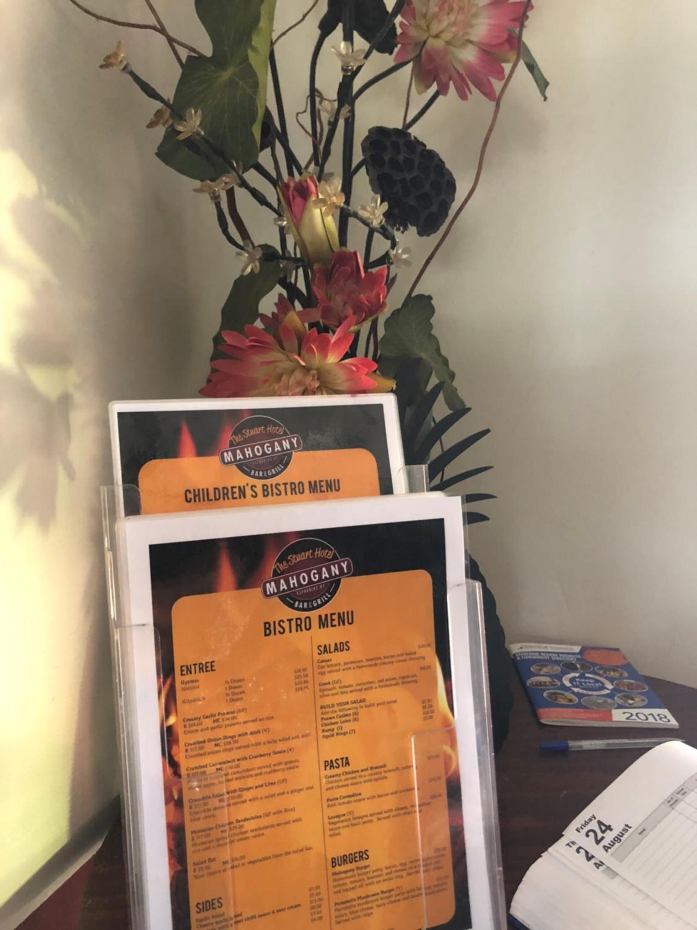 Cardápio de restaurante com área infantil, sobre uma mesa com um vaso de flores e um calendário.