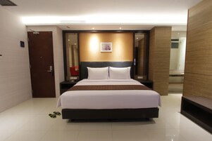Quarto de hotel com uma cama grande, mesa de cabeceira, cabeceira de madeira e porta para um banheiro.